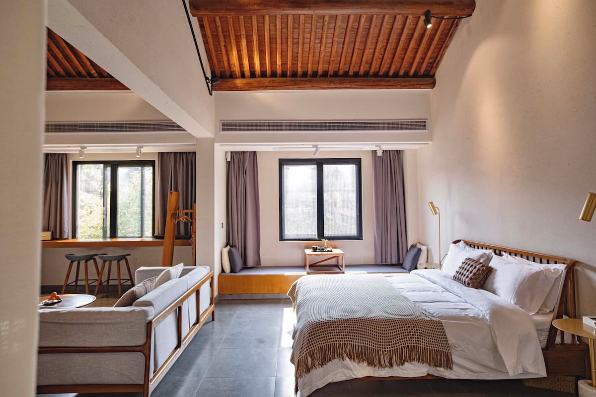 Quarto de hotel moderno com uma cama grande, um sofá, uma mesa pequena e um assento junto à janela.