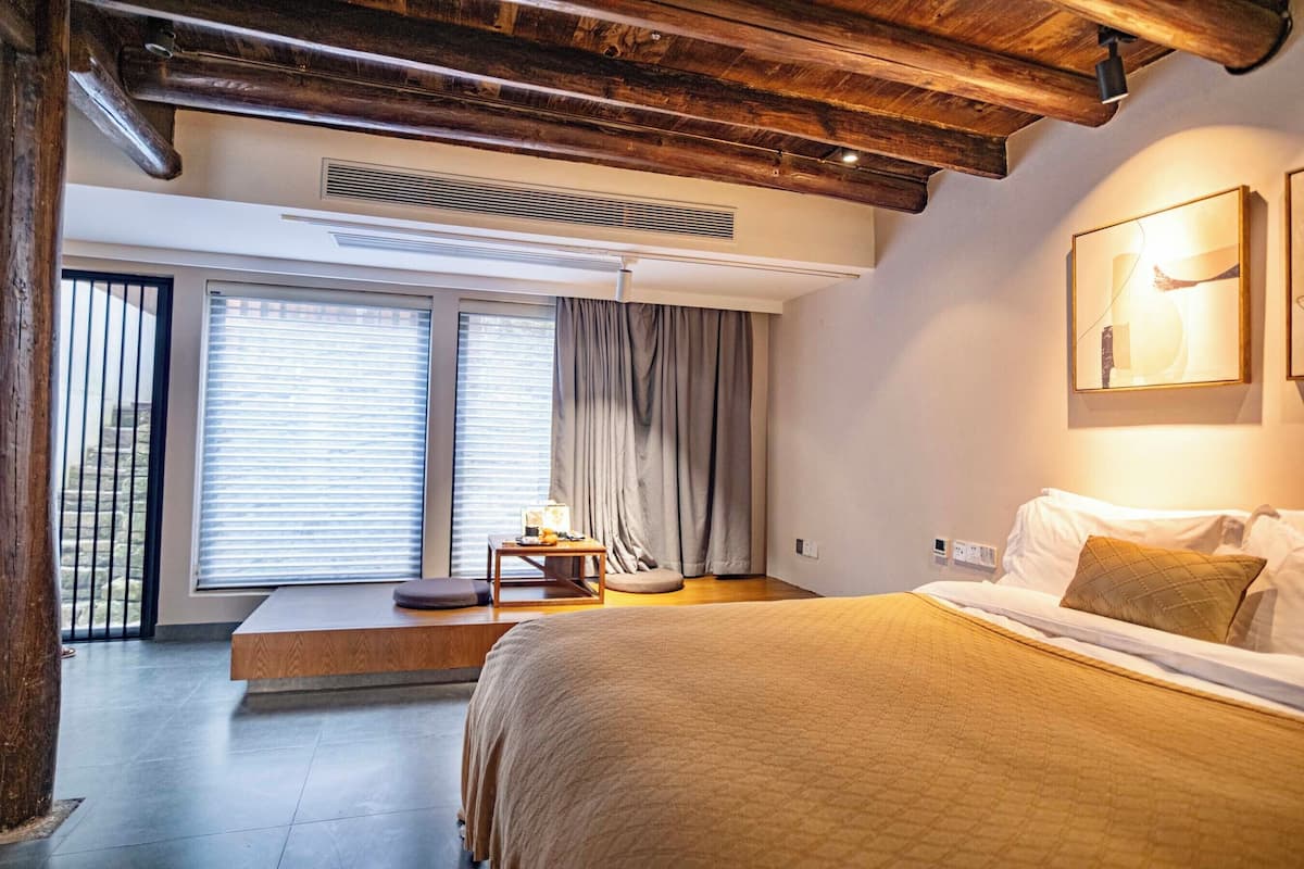 Quarto com cama, mesa de cabeceira, janela com cortinas, um banco de madeira e teto com vigas aparentes.