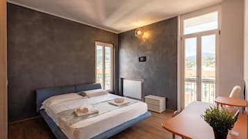 Quarto com cama, mesa de cabeceira e vista da cidade através de grandes janelas.
