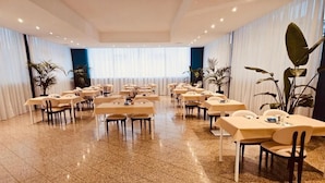 Sala de conferências com várias mesas e cadeiras, amplas janelas com cortinas e plantas em vasos.