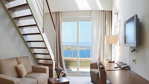 Quarto de hotel com sofá, mesa pequena com um vaso, escrivaninha com telefone, televisão e escada que leva a uma varanda com vista para o mar.