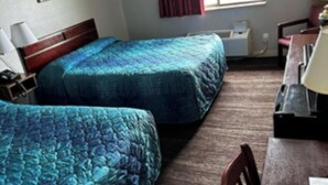 Quarto de hotel com duas camas, uma escrivaninha de madeira, uma cadeira, um abajur, uma janela com cortinas e uma TV fixada na parede.