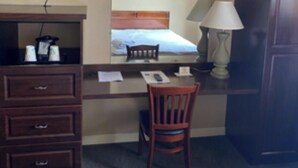 Quarto de hotel com cama, escrivaninha, cadeira, cômoda, abajur e televisão na parede.