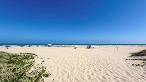 Uma extensa praia de areia dourada, céu azul límpido e mar calmo.