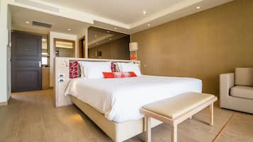 Quarto de hotel com uma cama grande, um banquinho, uma cadeira e um espelho.