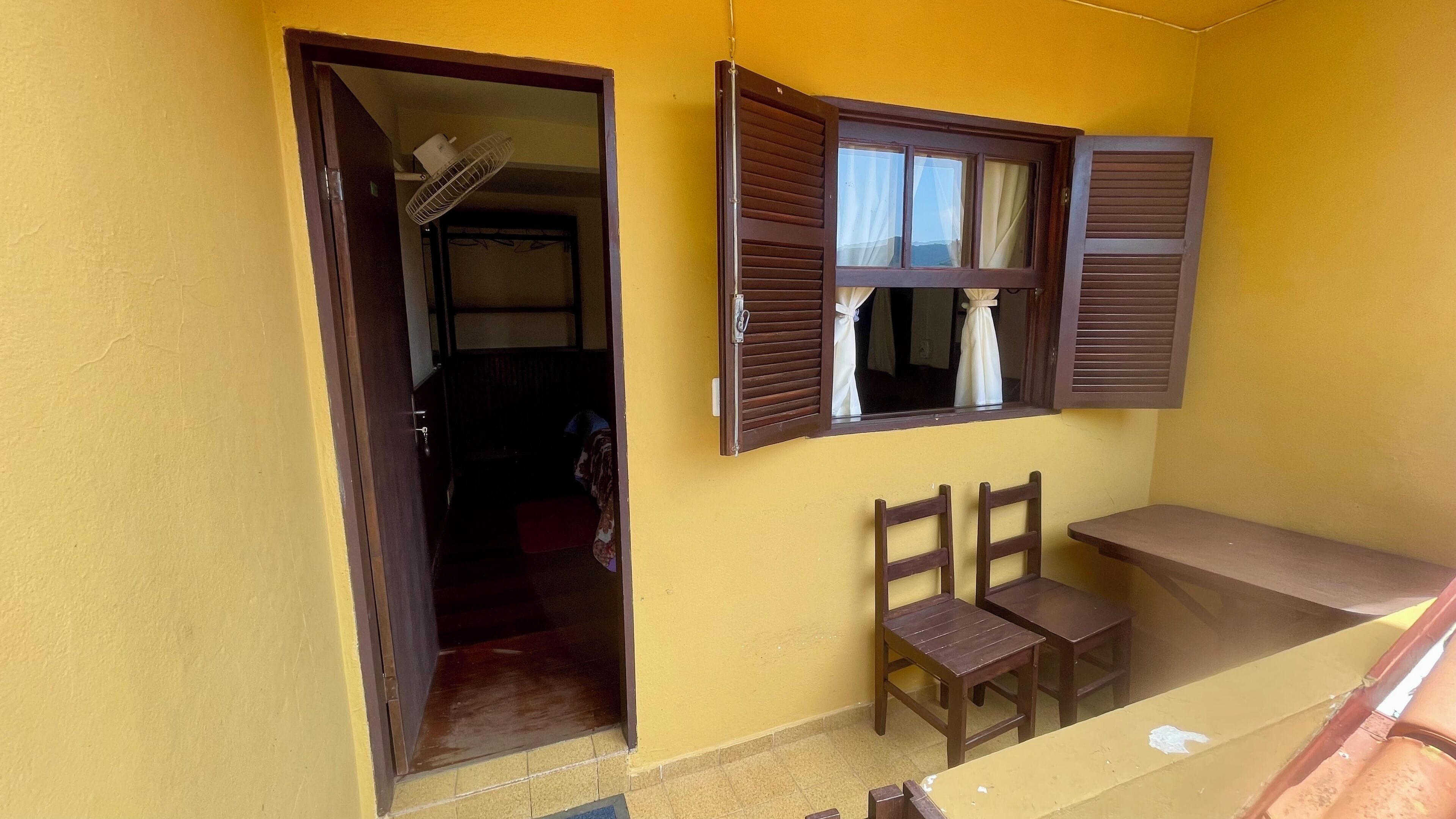 Um quarto com porta de madeira, janela com persianas, um ventilador e duas cadeiras de madeira.