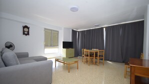 Uma sala de estar com um sofá, uma mesa de centro, uma mesa de jantar com cadeiras, uma televisão e um relógio na parede.