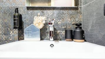 Casa de banho com uma pia branca, um espelho, uma embalagem de gel de banho, um dispensador de sabonete e um vaso decorativo.