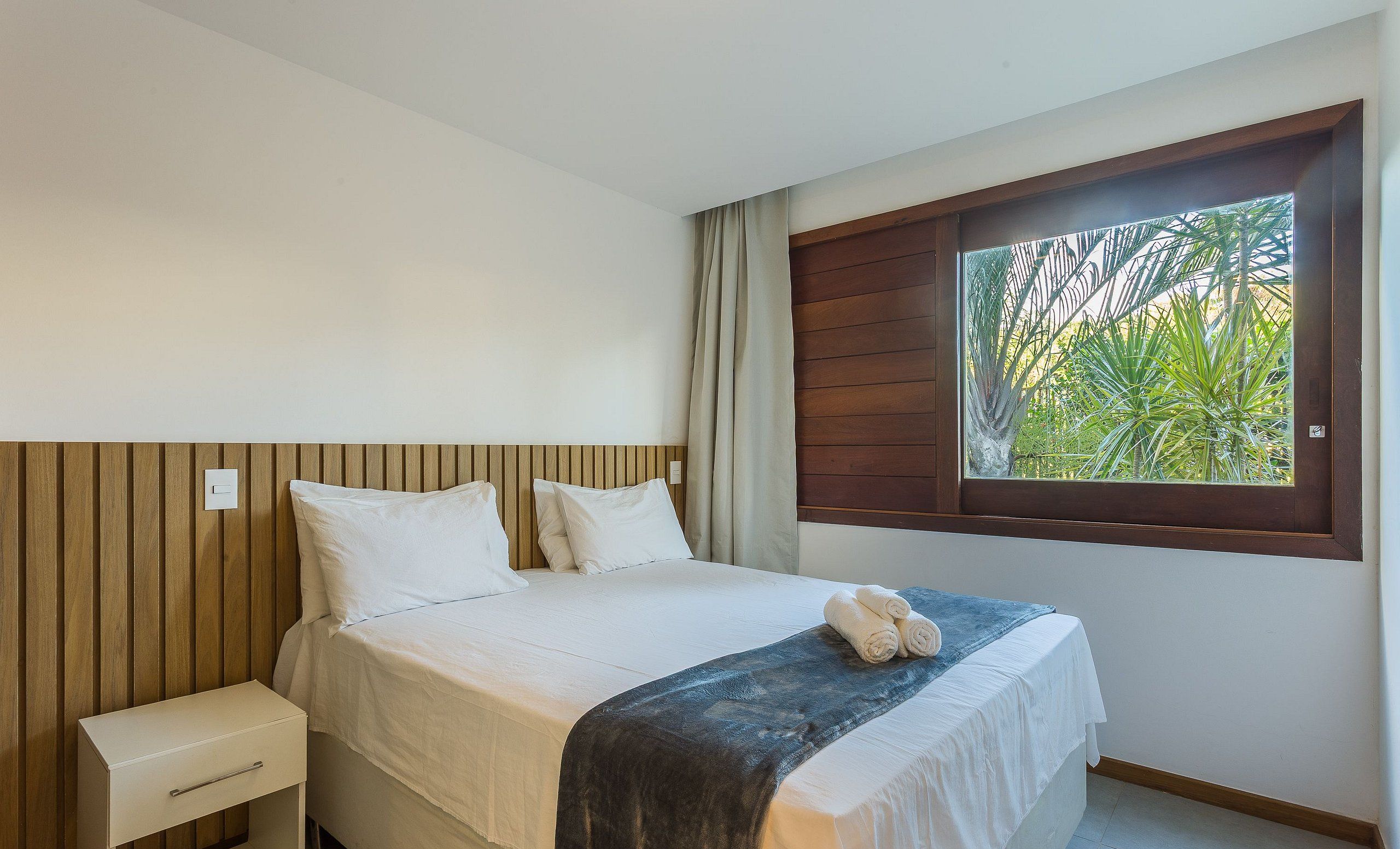Quarto com cama, mesa de cabeceira, janela com vista para palmeiras e cabeceira de madeira.