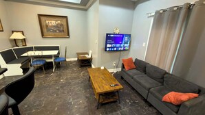 Sala de estar com um sofá preto, uma mesa de centro de madeira, uma cadeira azul, um sofá branco, uma TV fixada na parede e um quadro na parede.