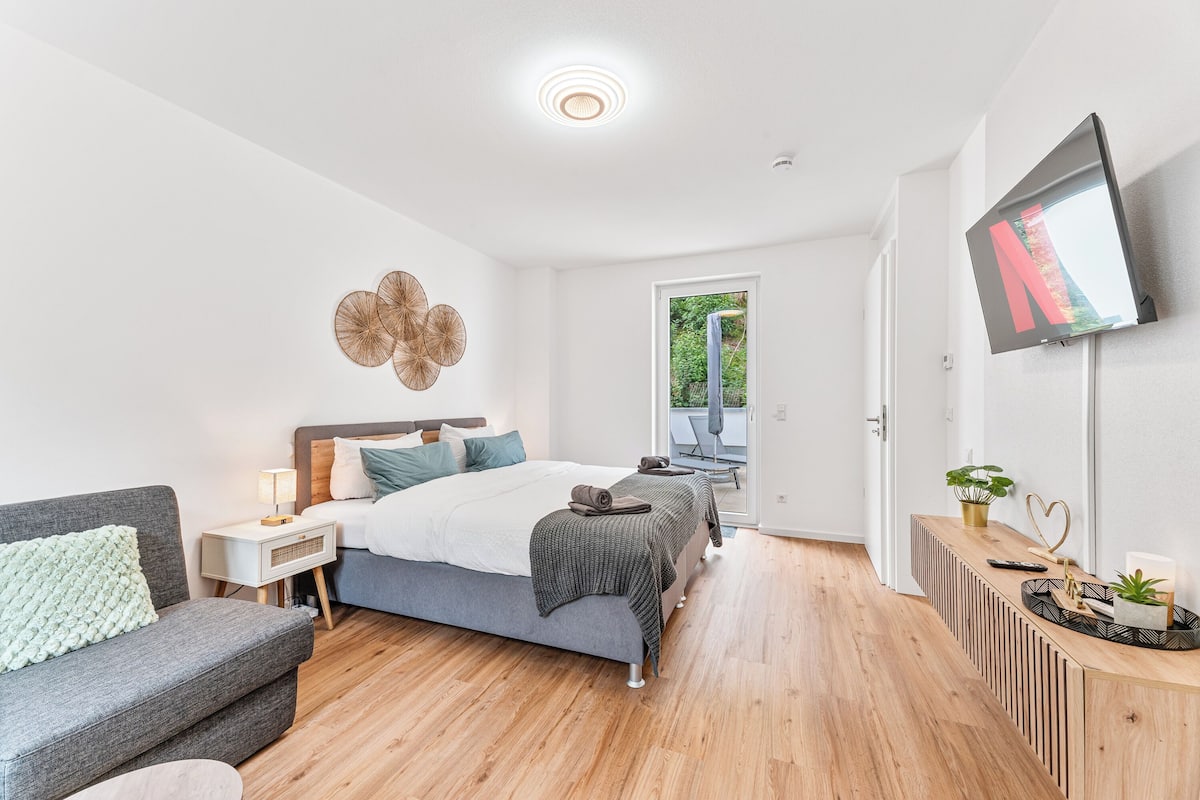 Quarto moderno com cama, sofá, televisão e uma pequena mesa com plantas.