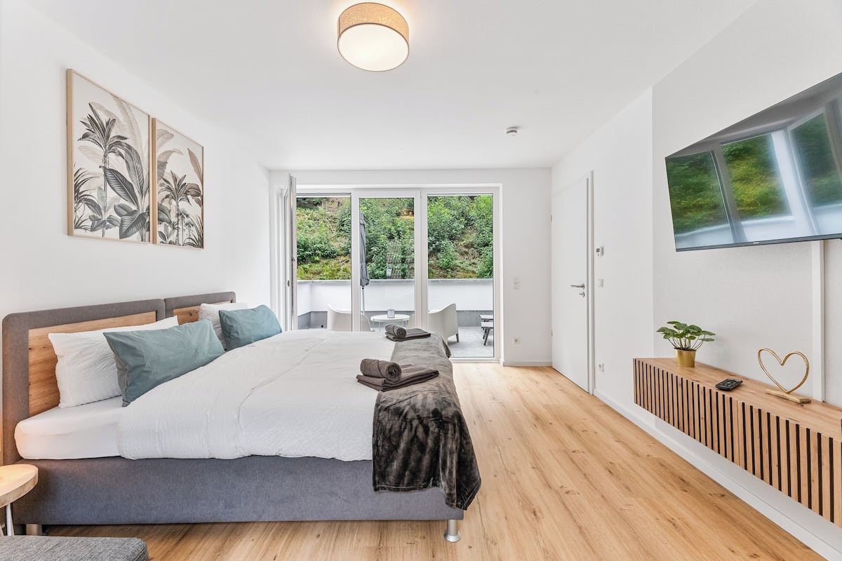 Quarto moderno com uma cama grande, uma televisão e uma varanda com vista para o verde.