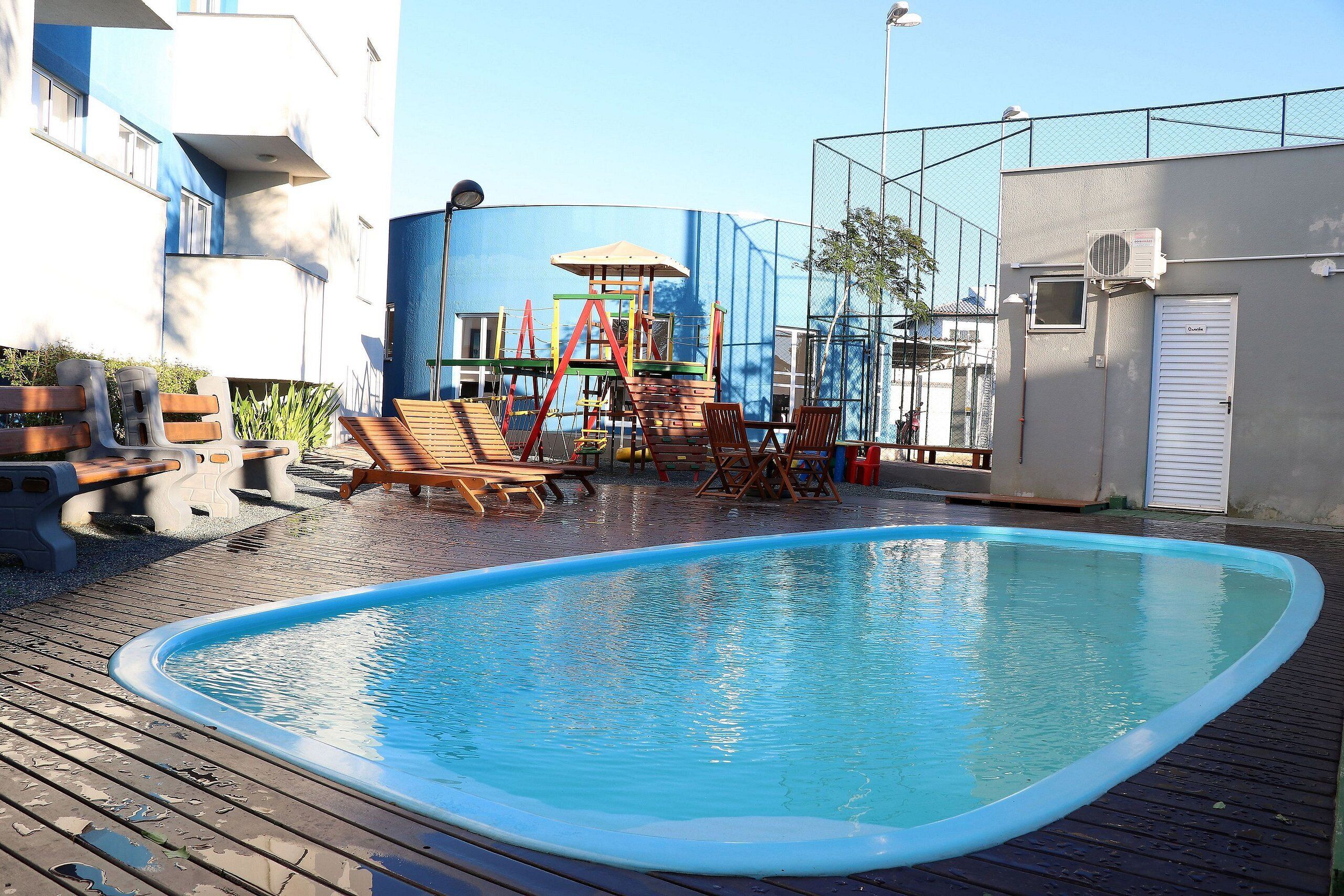 Uma piscina com deck de madeira, espreguiçadeiras e um parquinho infantil.