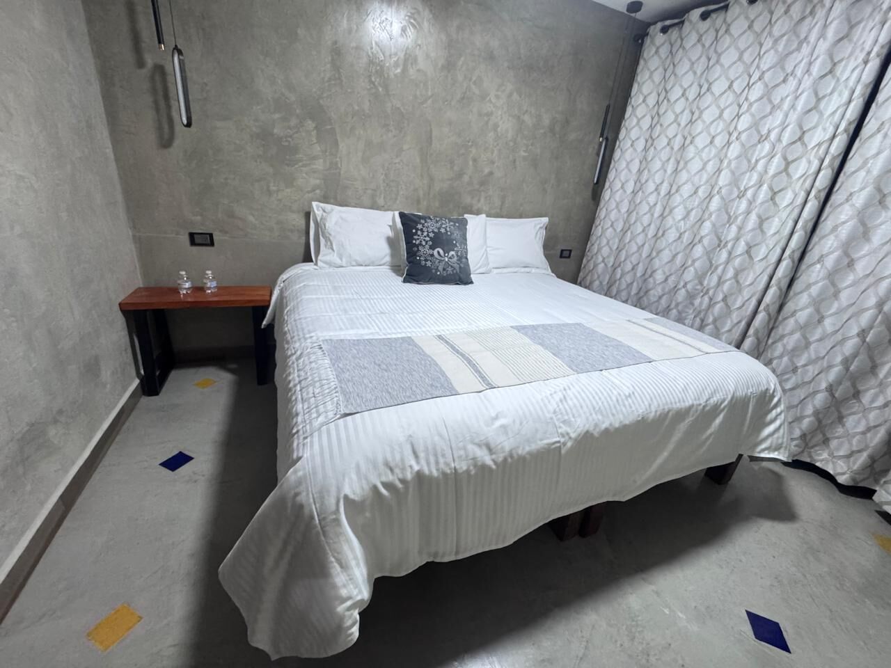 Um quarto com cama, mesa de cabeceira e cortina.