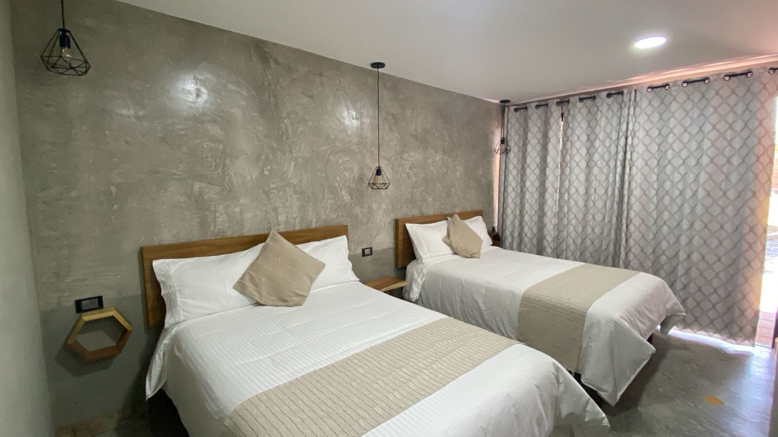 Quarto de hotel com duas camas, uma parede de concreto, luminária pendente e uma janela com cortinas.