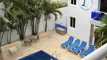 Piscina com espreguiçadeiras, uma mesa pequena e cadeiras, cercada por palmeiras.