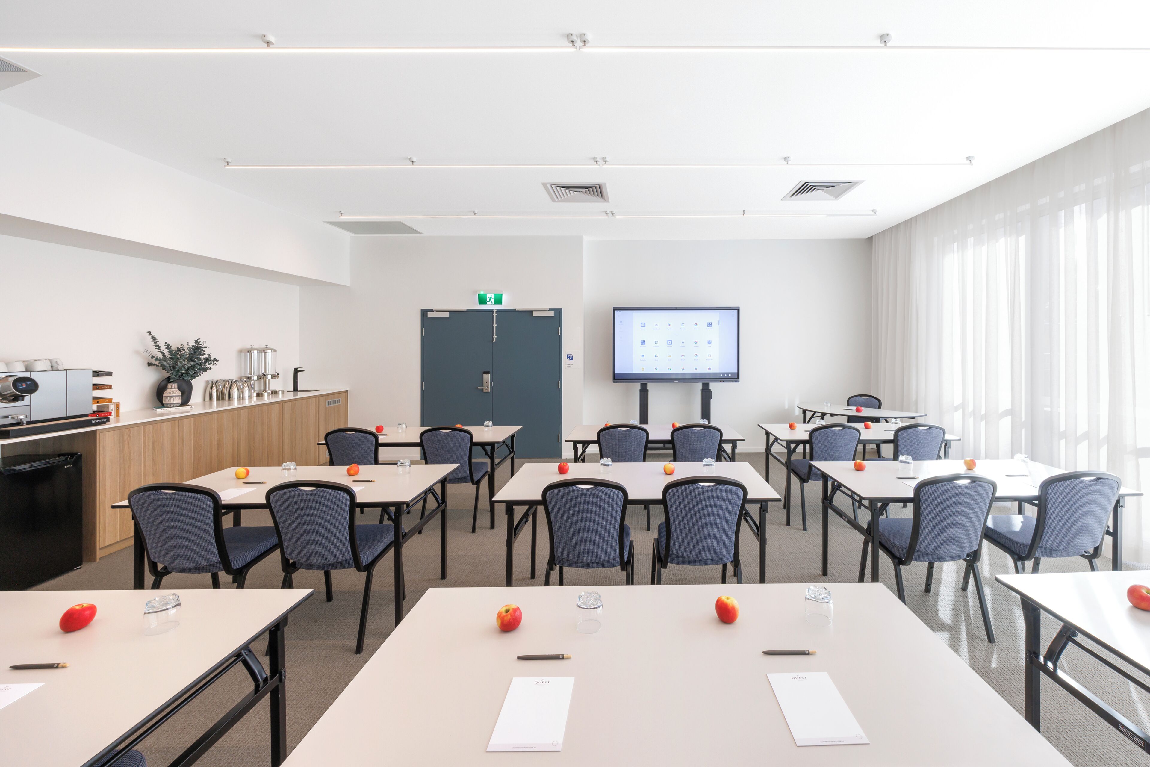 Sala de conferências com mesas, cadeiras, tela de projetor e uma pequena copa.