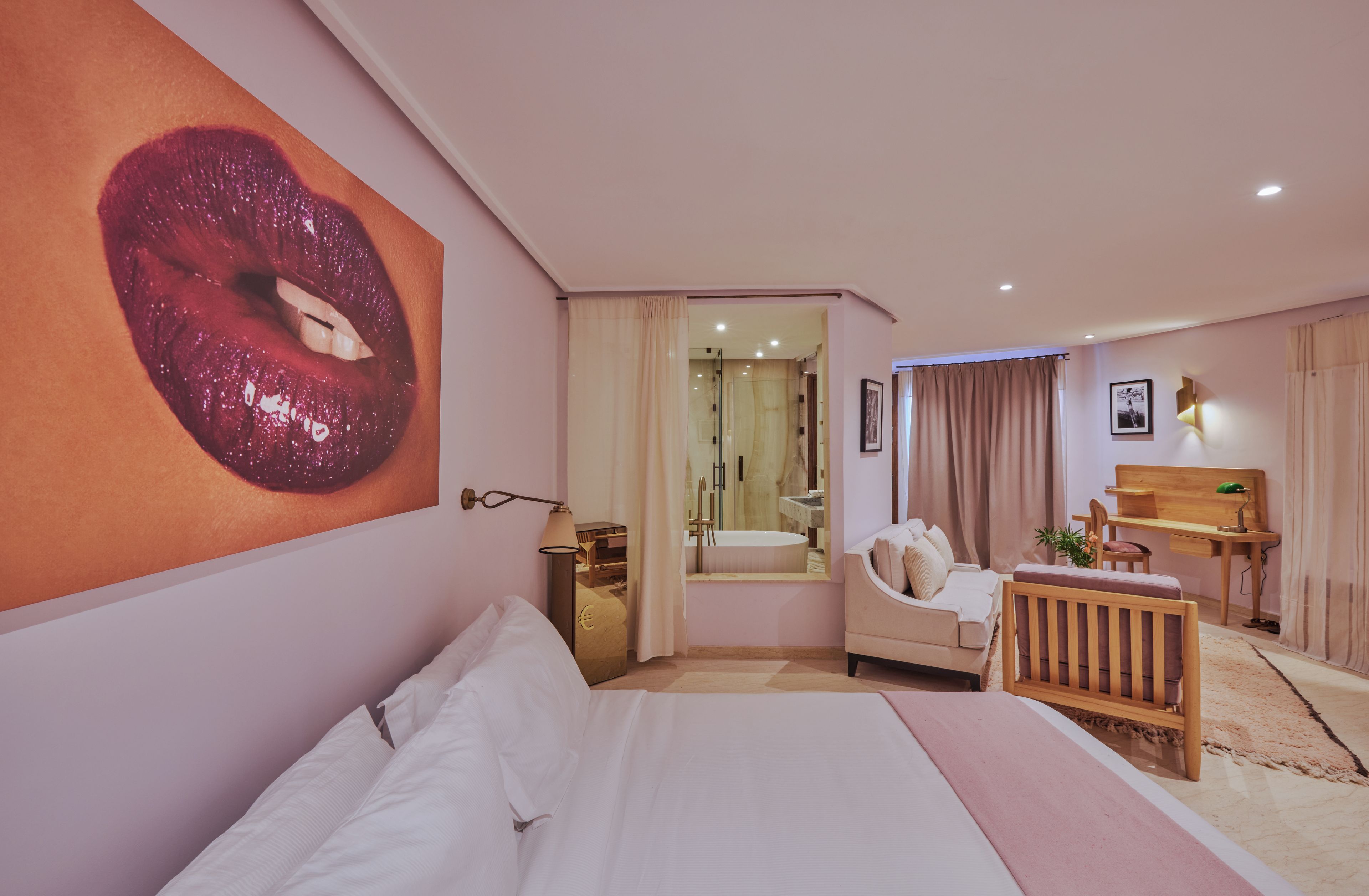 Quarto de hotel moderno com uma cama grande, área de estar com sofá e uma mesa pequena, um espelho grande e um banheiro visível através de uma porta aberta.