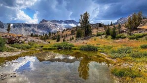 Paisagem montanhosa com um lago límpido refletindo o céu e árvores ao redor.
