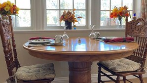 Uma sala de jantar com uma mesa redonda de madeira, quatro cadeiras e uma grande janela com cortinas. Há arranjos florais no parapeito da janela e um vaso sobre a mesa.