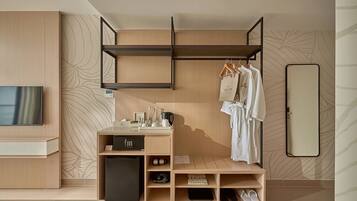 Quarto de hotel moderno com televisão, frigobar, escrivaninha com cafeteira, guarda-roupa com roupas penduradas e espelho.