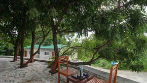 Uma área de estar coberta ao ar livre com duas cadeiras de madeira e uma mesa, cercada por árvores e um edifício com telhado verde ao fundo.