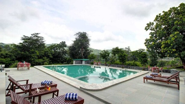 Piscina retangular com espreguiçadeiras e mesas ao redor, cercada por árvores e colinas.