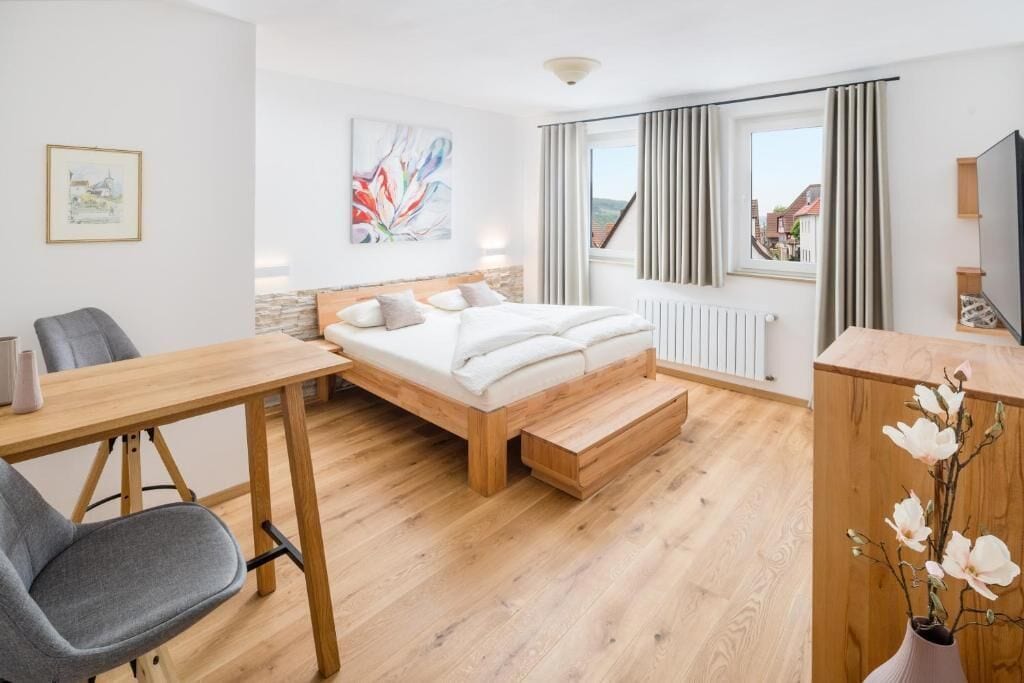 Quarto moderno com cama, escrivaninha e cadeira, janela com cortinas e vista para edifícios.