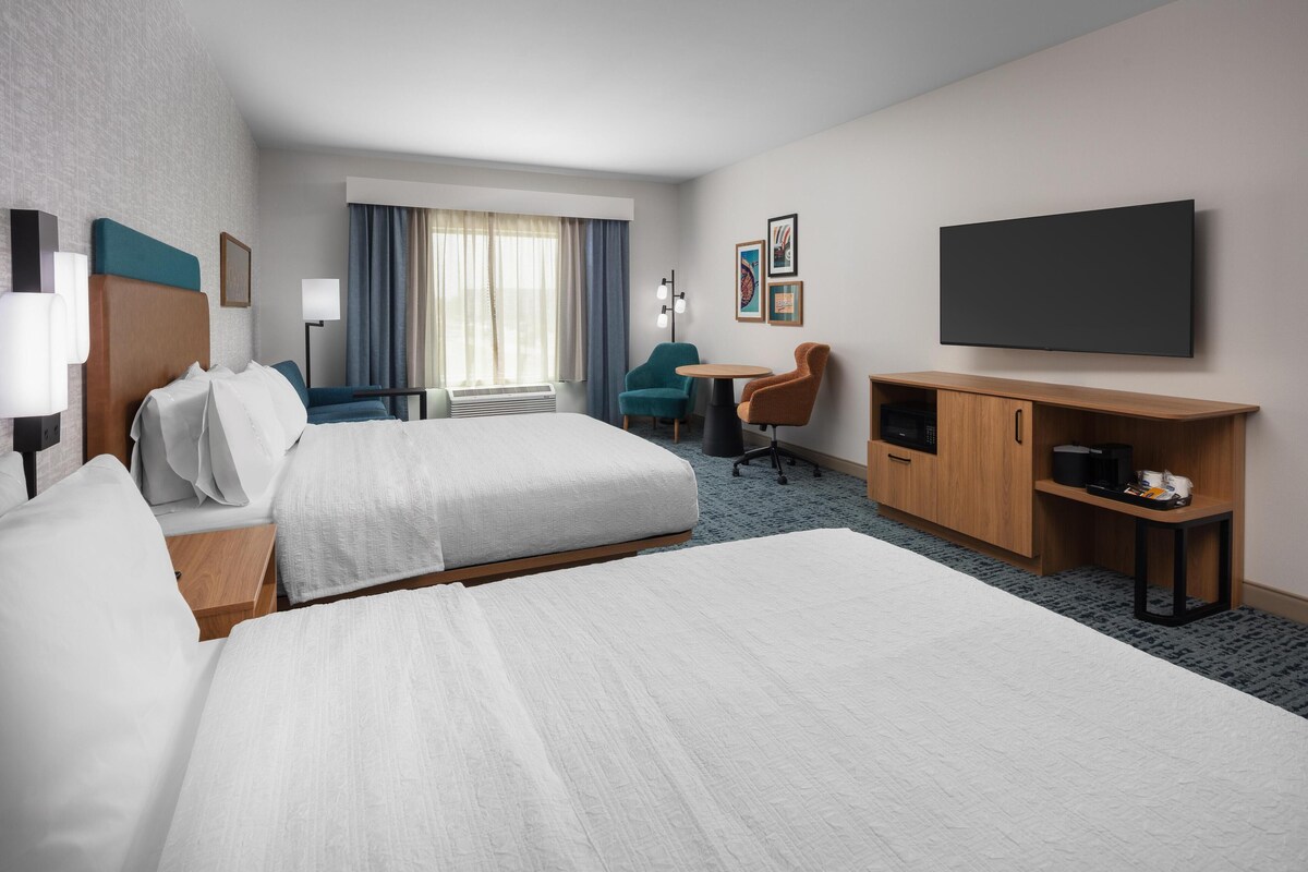 Quarto de hotel com duas camas, uma escrivaninha, uma cadeira, uma televisão e uma janela com cortinas.