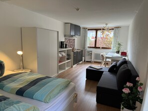 Quarto com cama, luminária de cabeceira, armário, cozinha compacta e área de estar com sofá e mesa.