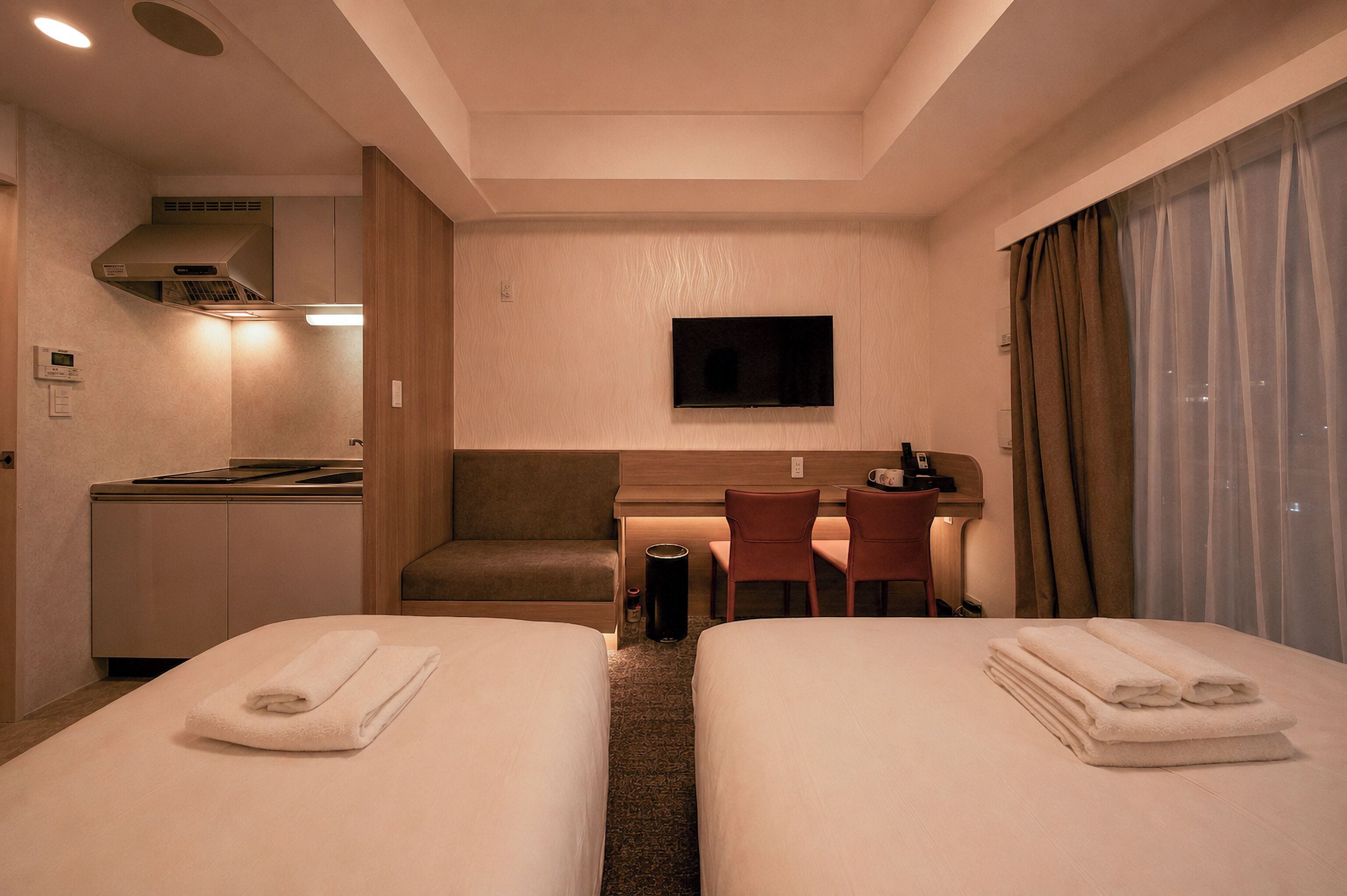 Quarto de hotel com duas camas, uma escrivaninha, uma cadeira, uma televisão e uma janela com cortinas.