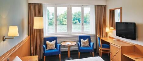 Quarto de hotel com cama, duas cadeiras azuis, uma mesa pequena, escrivaninha, televisão e janela com vista para um corpo de água.