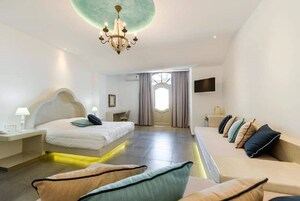Quarto de hotel moderno com cama de casal, sofá, televisão e lustre.