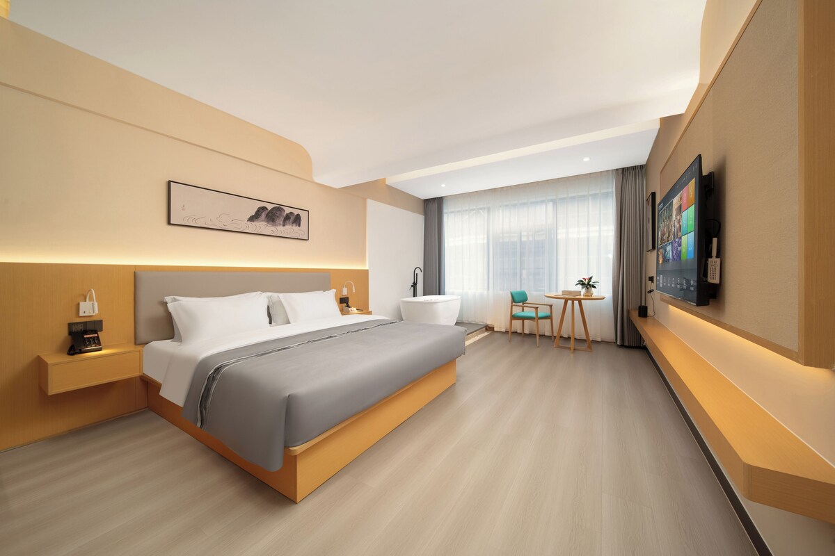 Quarto de hotel moderno com cama grande, mesa de cabeceira, televisão fixada na parede e um pequeno espaço de estar com mesa e cadeira.