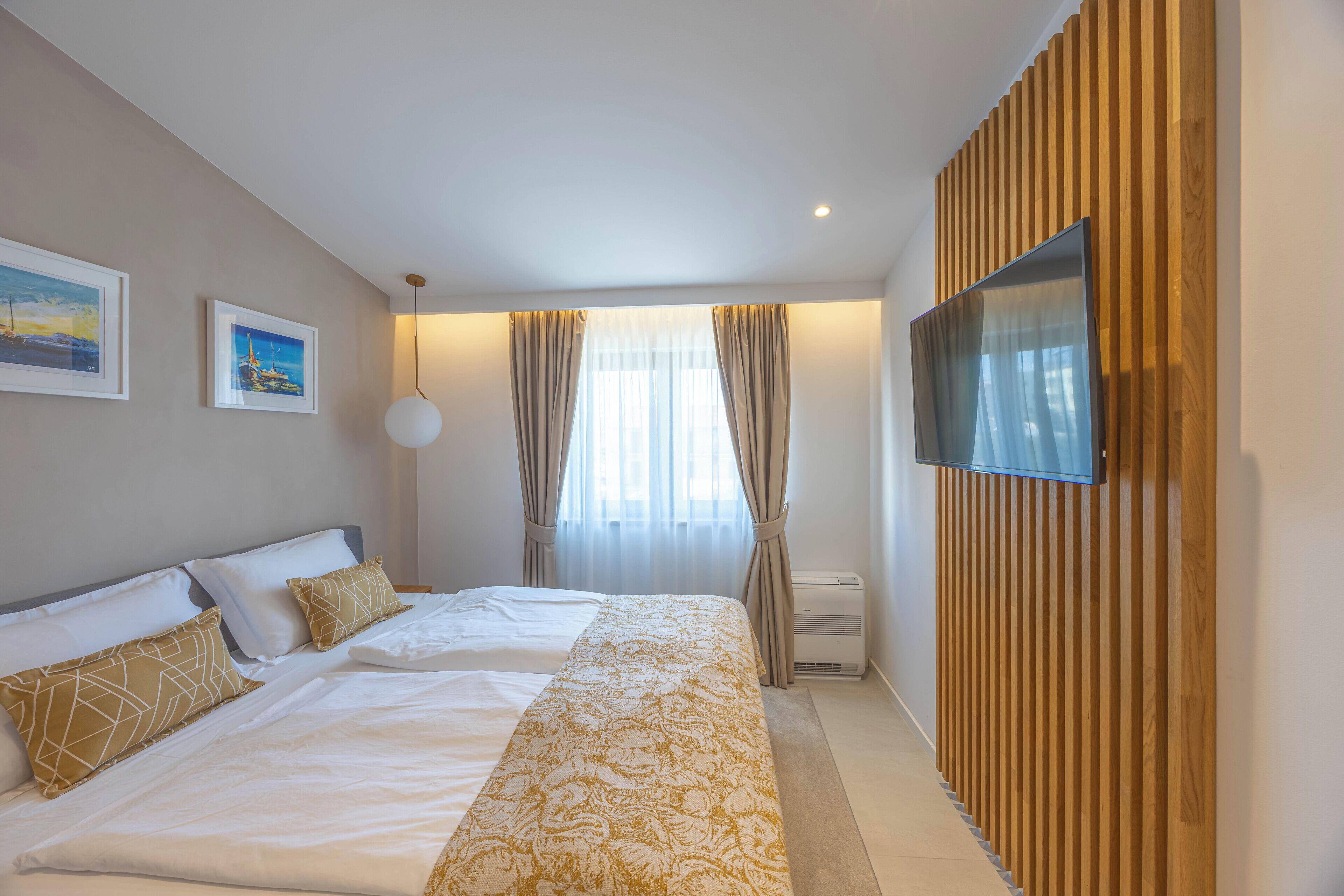 Quarto de hotel com cama, televisão fixada em um painel de madeira e janela com cortinas.