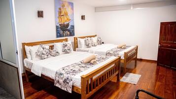Quarto de hotel com duas camas de madeira, um banco de madeira, um ventilador de teto e um quadro de um navio na parede.