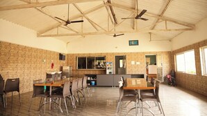 Um quarto amplo com teto alto, vigas aparentes e ventoinhas. Há mesas e cadeiras dispostas para refeições, uma cozinha equipada e uma parede de tijolos.