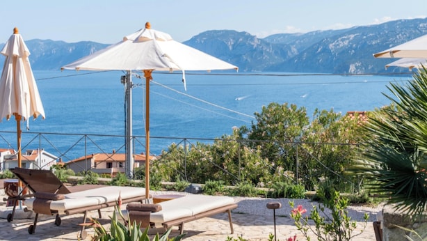 Terraço à beira-mar com espreguiçadeiras e guarda-sóis, com vista para um mar azul cristalino e montanhas ao longe.