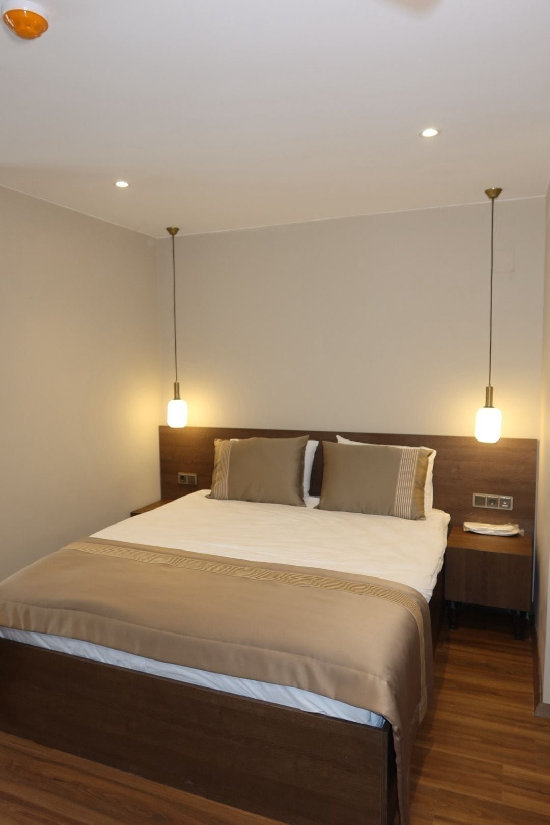 Quarto de hotel com uma cama grande, duas mesas de cabeceira, uma cabeceira de madeira e luminárias pendentes.