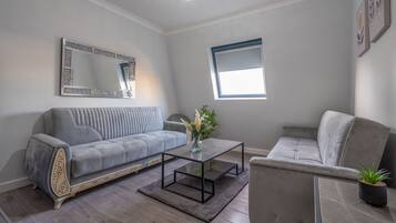Uma sala de estar moderna com um sofá cinzento, uma mesa de centro de vidro e uma poltrona cinzenta. Um espelho grande está pendurado na parede, e há uma janela com moldura azul.