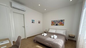 Quarto de hotel com uma cama, uma cadeira, uma escrivaninha, aparelho de ar condicionado e um quadro na parede.