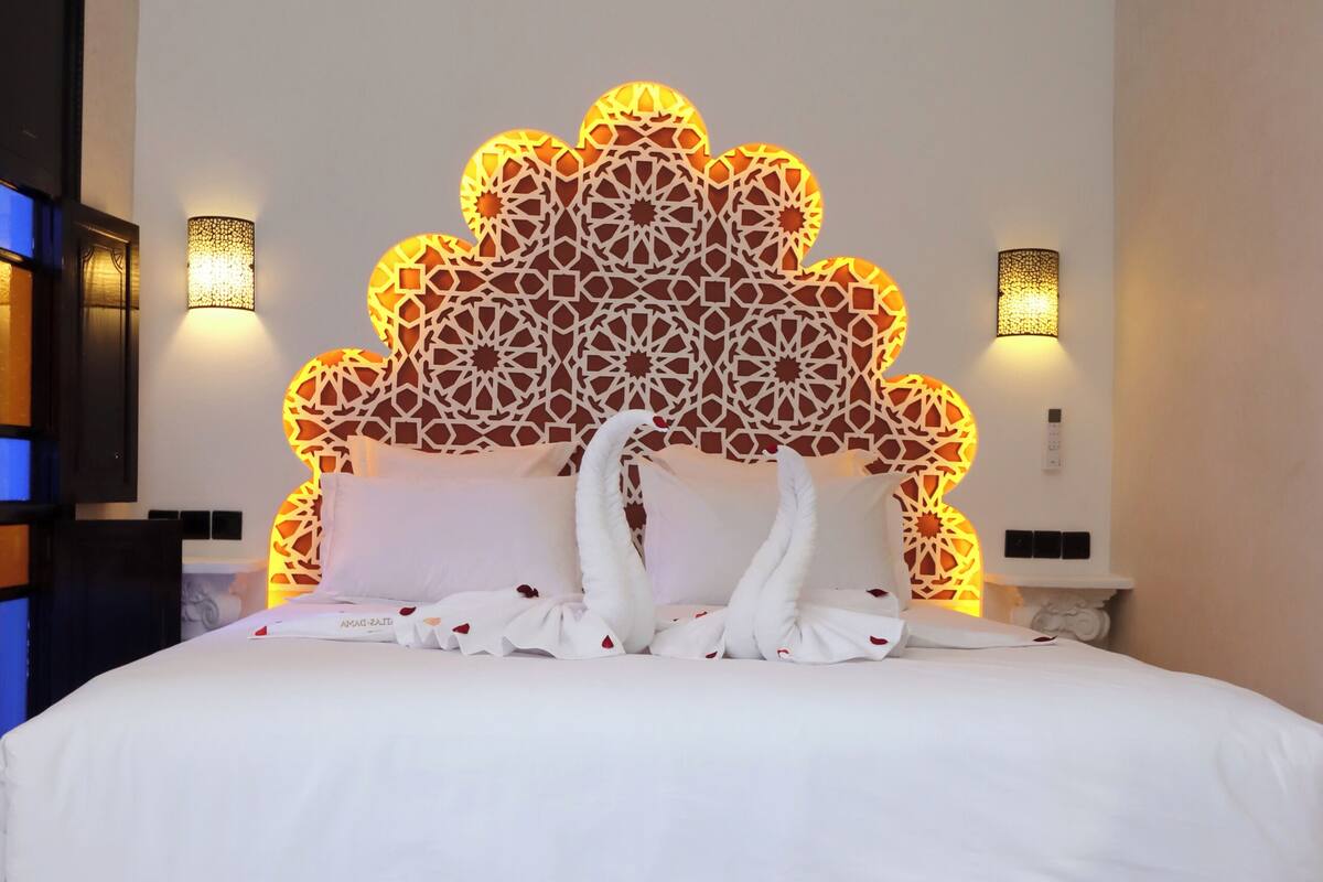 Quarto de hotel com uma cama grande, cabeceira decorativa, arandelas de parede e tapete estampado.