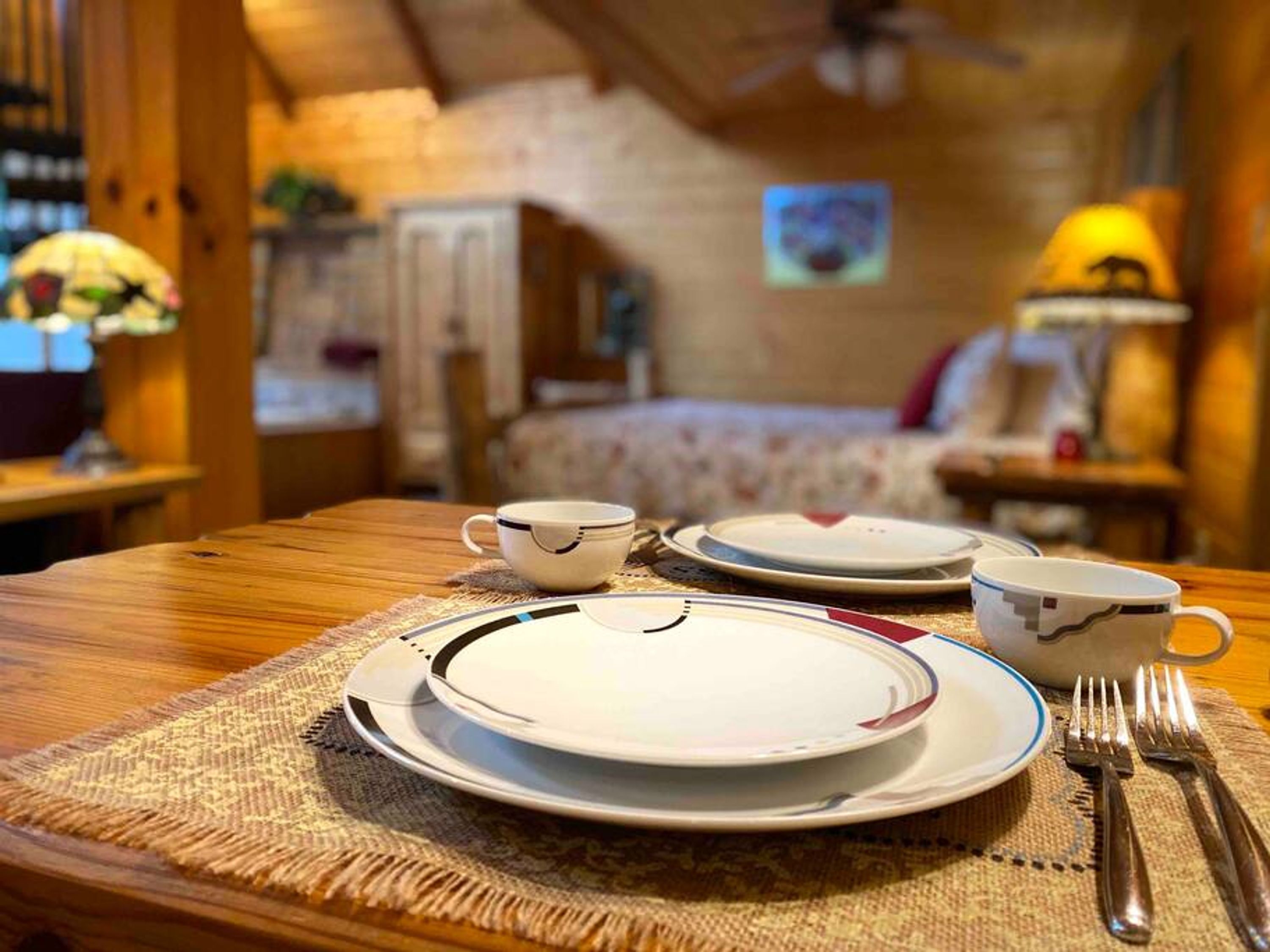 Uma mesa de jantar de madeira com pratos, copos e talheres, montada em um ambiente acolhedor com uma cama e um abajur ao fundo.