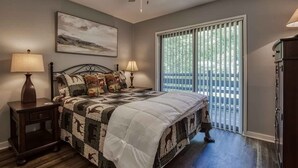 Quarto com cama, duas luminárias de cabeceira, ventilador de teto, janela com persianas e vista para um deck.