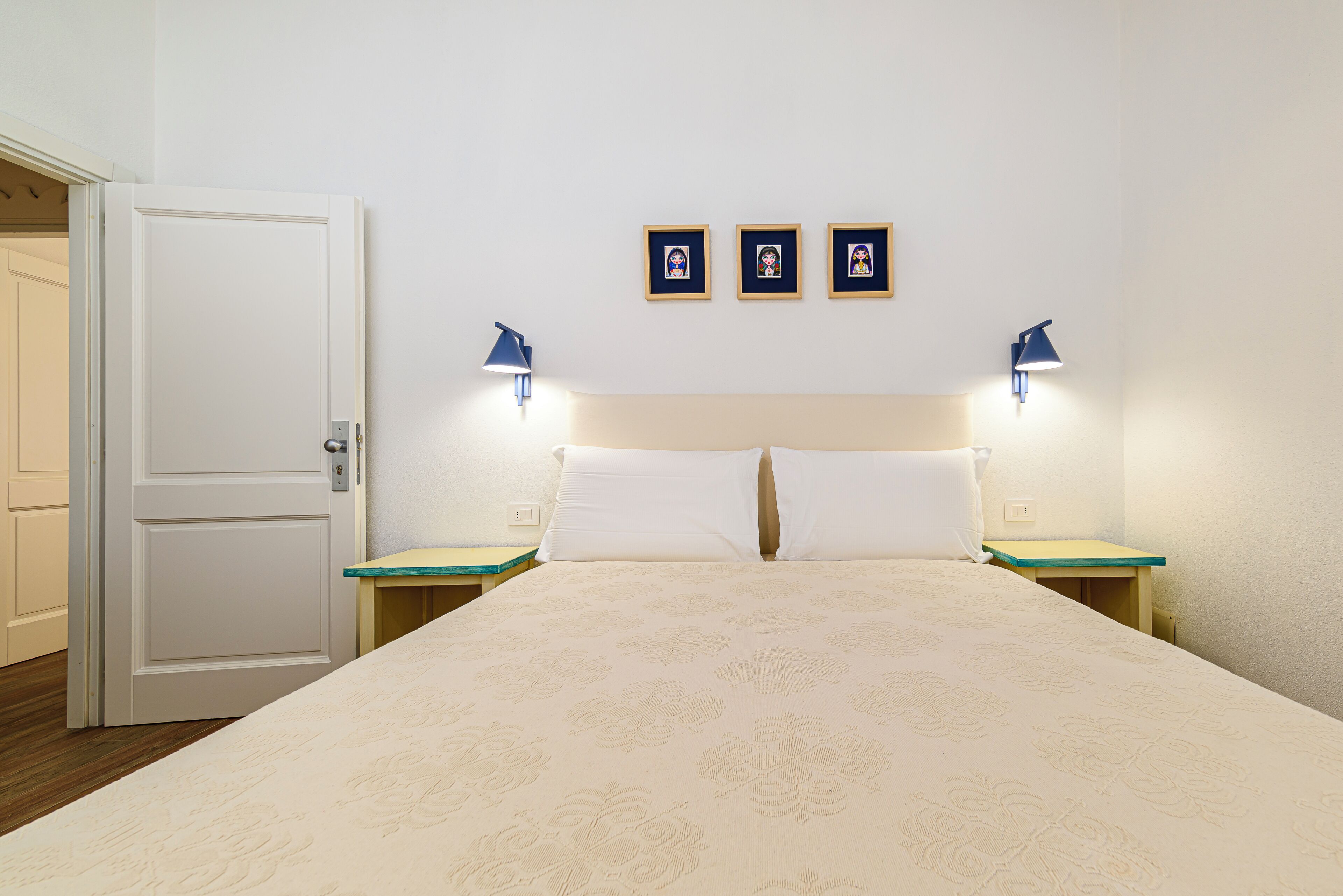 Quarto de hotel com uma cama, duas mesinhas de cabeceira, um criado-mudo e três quadros na parede.