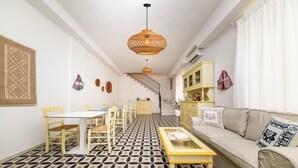 Uma sala de estar com piso estampado, um sofá, uma mesa de centro, uma mesa de jantar com cadeiras, uma escada e um lustre.