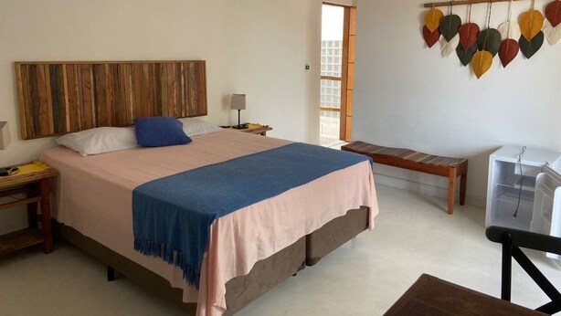 Quarto com uma cama grande, cabeceira de madeira, um banquinho, uma mesa pequena e um objeto decorativo na parede.
