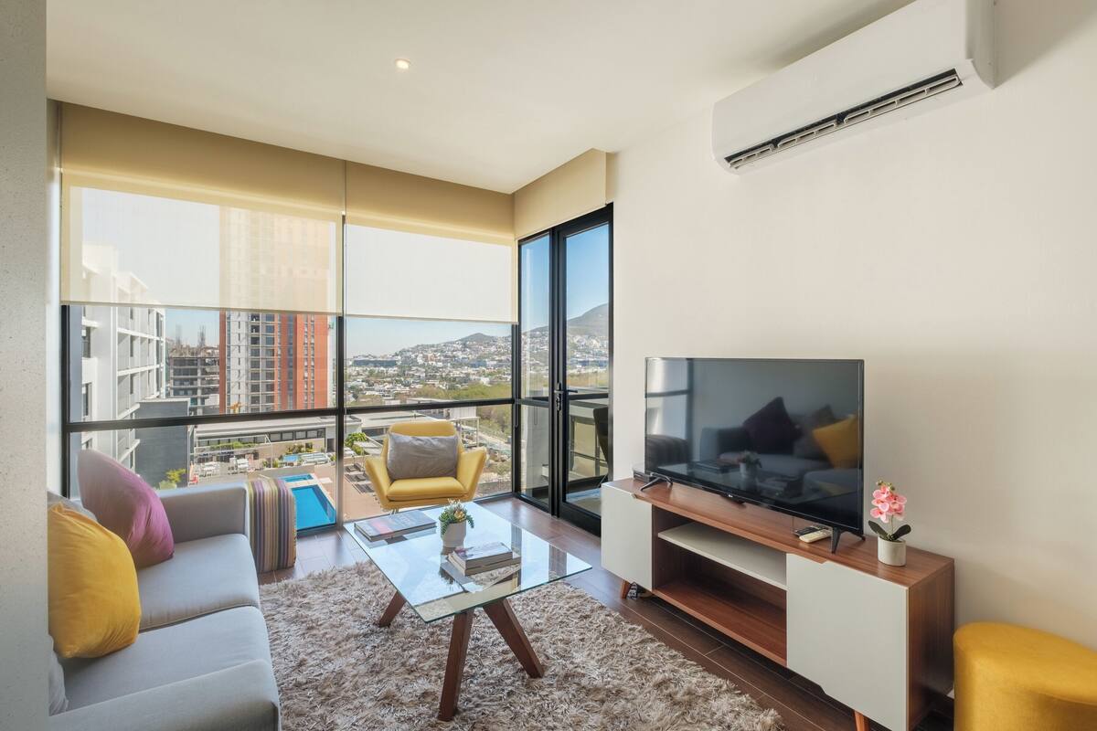 Uma sala de estar moderna com sofá, mesa de centro, TV e uma grande janela com vista para a cidade.