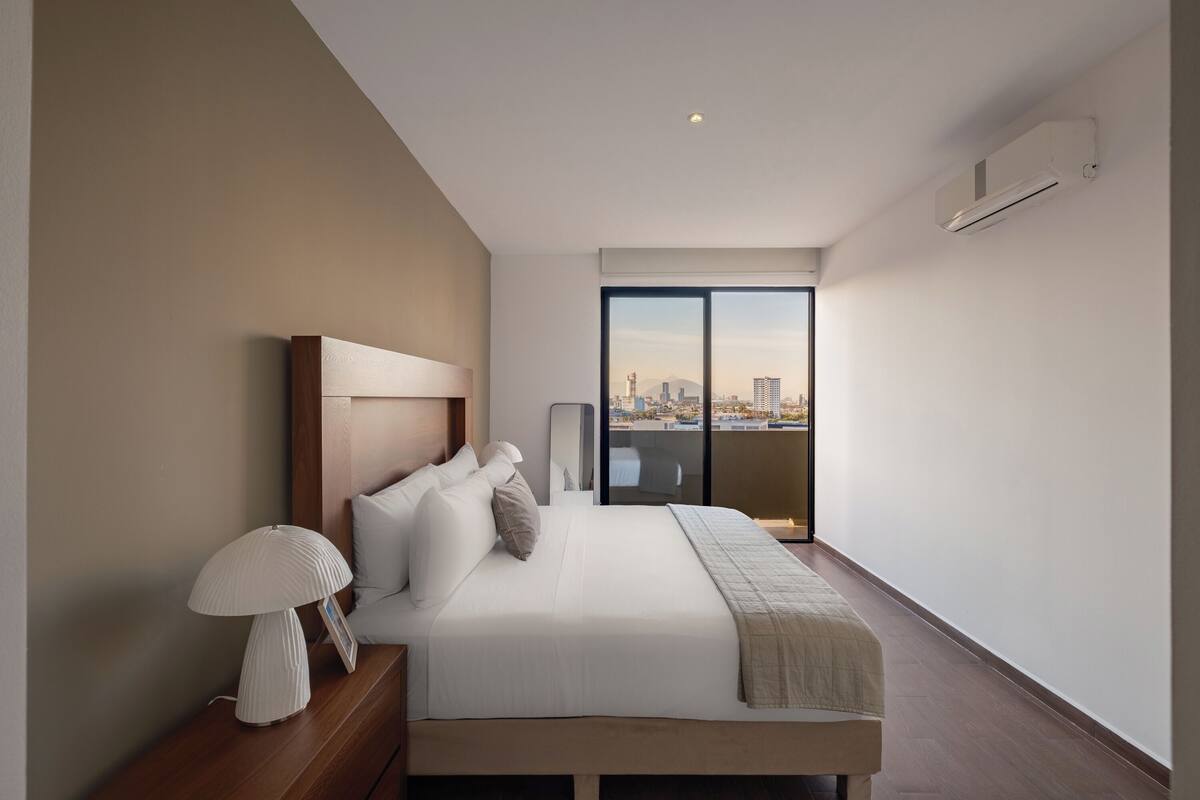 Quarto de hotel moderno com uma cama grande, abajur, televisão e vista para a cidade através de uma janela ampla.