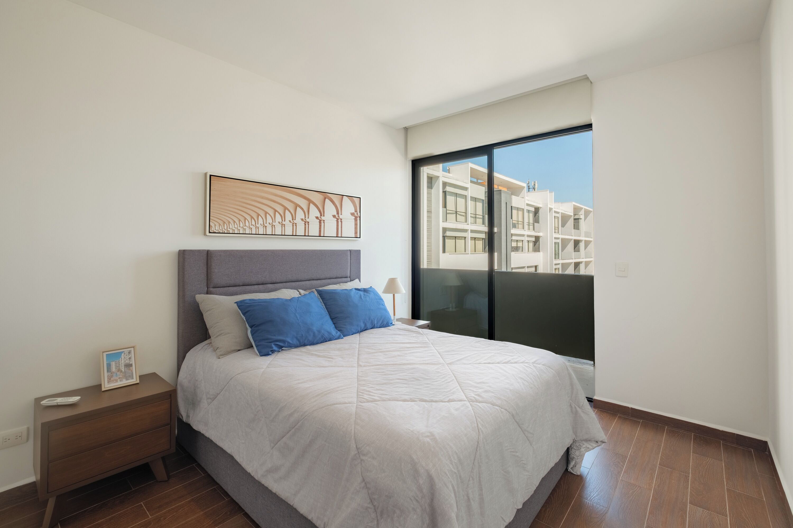 Quarto moderno com uma cama grande, uma mesinha de cabeceira, um quadro emoldurado e vista para edifícios através de uma janela ampla.